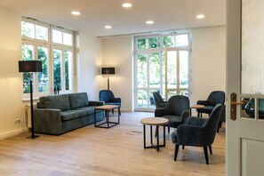 Quarto espaçoso com sofá cinzento, duas poltronas, uma mesinha e uma luminária de chão. Grandes janelas oferecem luz natural e vista para o verde lá fora.