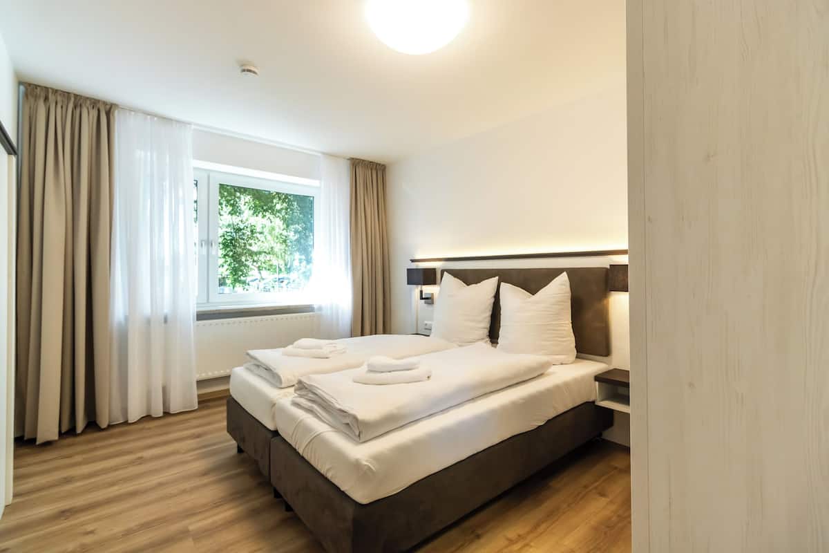 Quarto de hotel moderno com uma cama grande, uma mesa de cabeceira, uma janela com cortinas e vista para o verde lá fora.