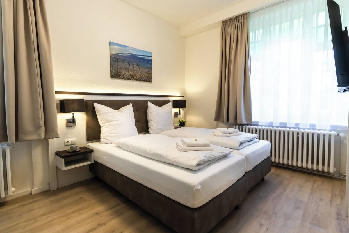 Uma cama de casal com roupa de cama branca, uma mesa de cabeceira com abajur, uma TV fixada na parede e uma janela com cortinas.