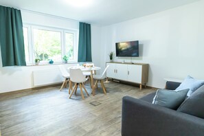Uma sala de estar moderna com área de jantar, um sofá cinzento, televisão de tela plana e uma janela com cortinas.