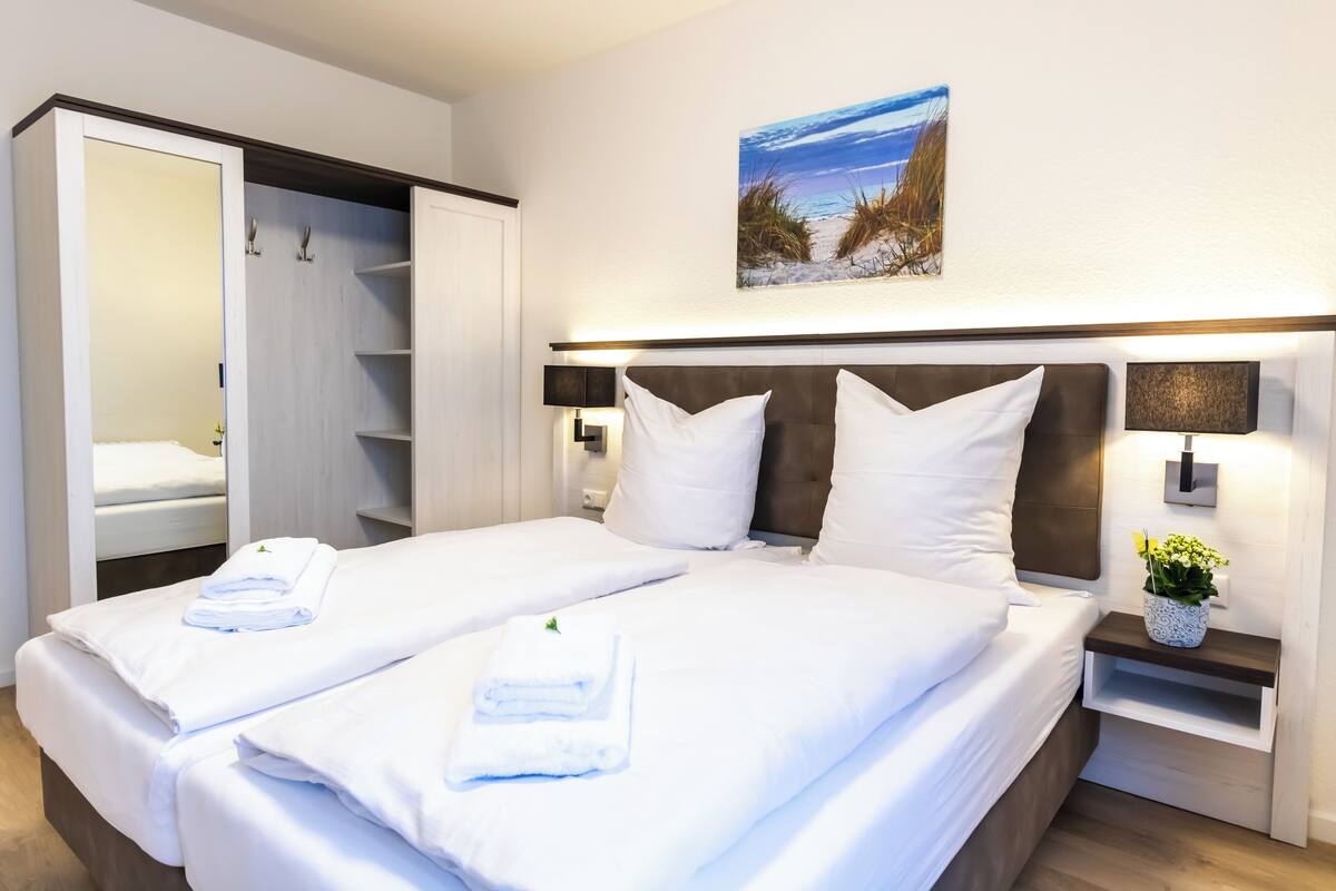 Quarto de hotel com uma cama, um guarda-roupa, uma mesa de cabeceira com uma planta e um quadro na parede.