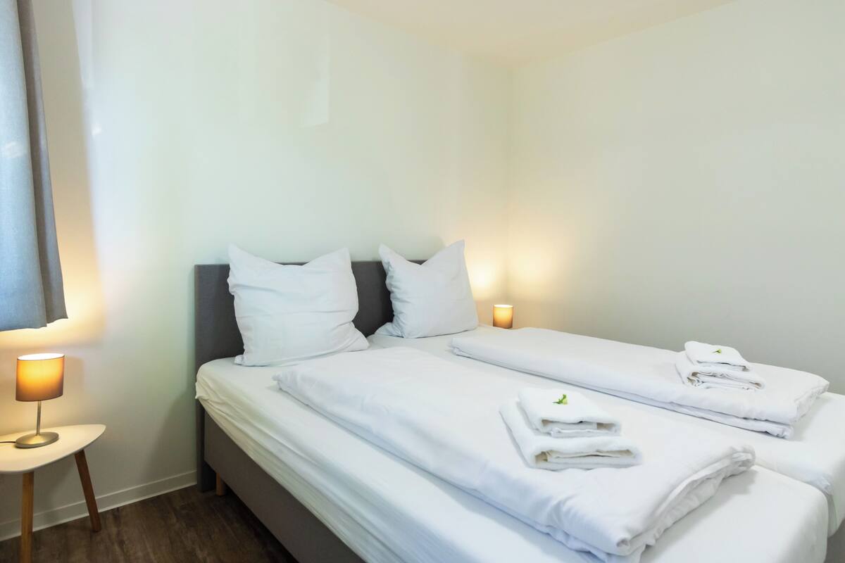 Quarto de hotel com duas camas, roupa de cama branca, mesa de cabeceira com abajur e criado-mudo.
