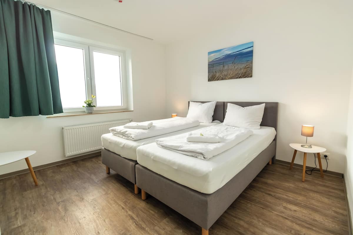 Uma cama de casal com roupa de cama branca, uma pequena mesa de cabeceira com abajur, uma janela com cortinas e um vaso de planta no parapeito.