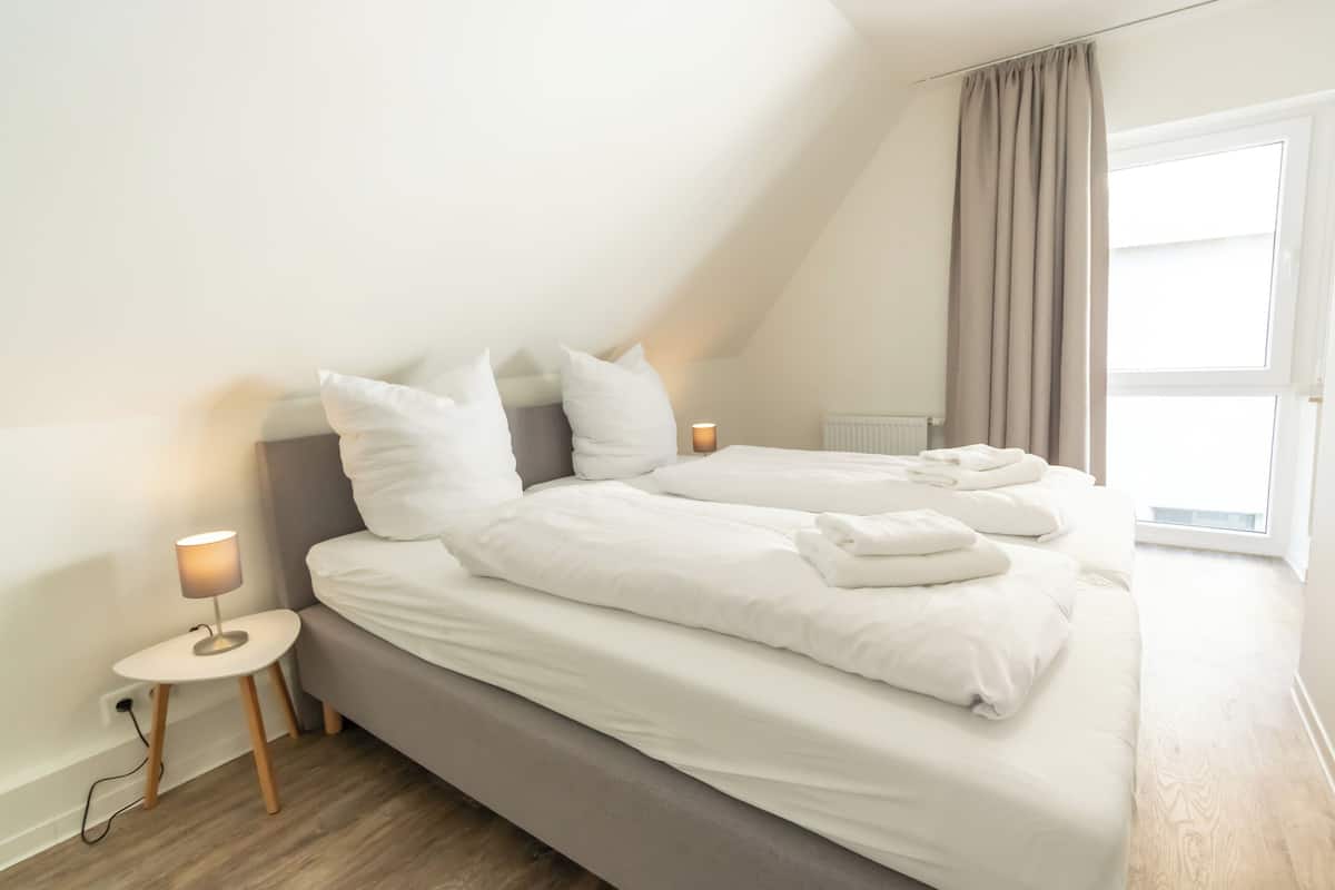 Quarto com duas camas, mesa de cabeceira com luminária, janela com cortinas e radiador.