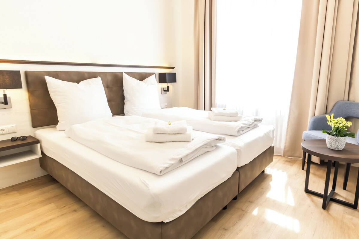Quarto de hotel com duas camas, uma mesinha com uma planta e uma mesa de cabeceira com abajur.