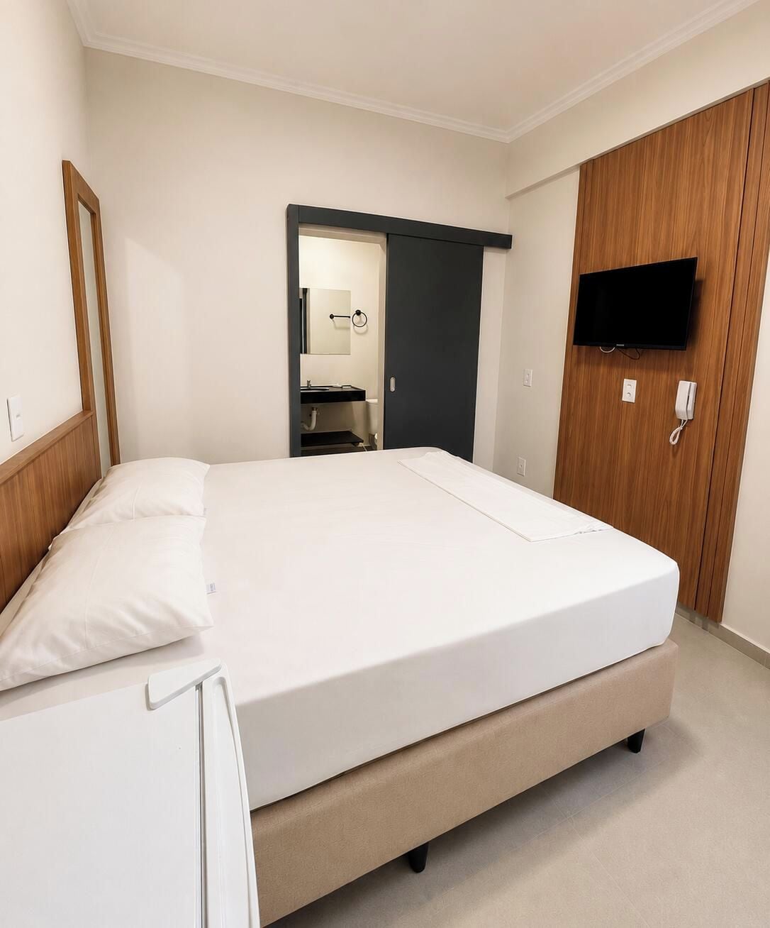 Quarto de hotel com cama, televisão fixada na parede, guarda-roupa de madeira e casa de banho visível através de uma porta aberta.