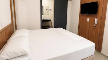 Quarto de hotel com cama, televisão fixada na parede, guarda-roupa de madeira e casa de banho visível através de uma porta aberta.
