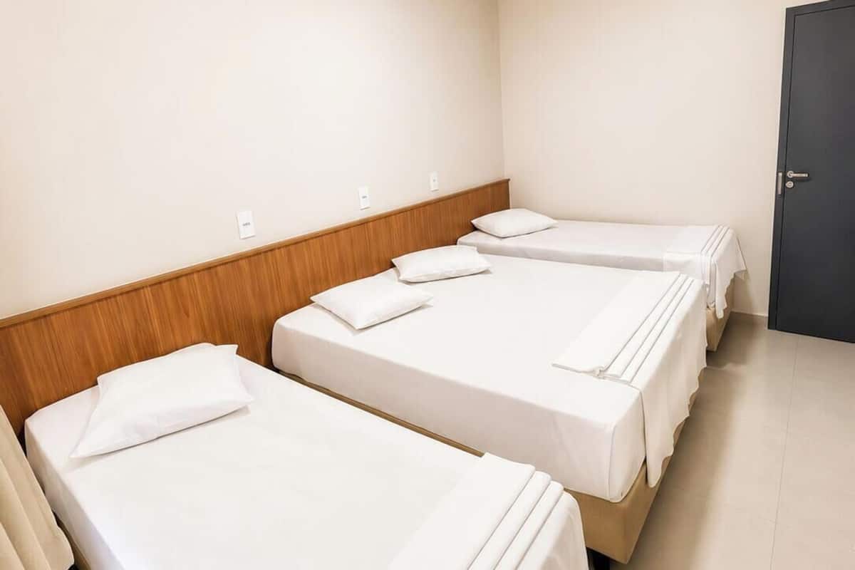 Quarto de hotel com três camas de solteiro, cabeceira de madeira, porta escura e chão ladrilhado.