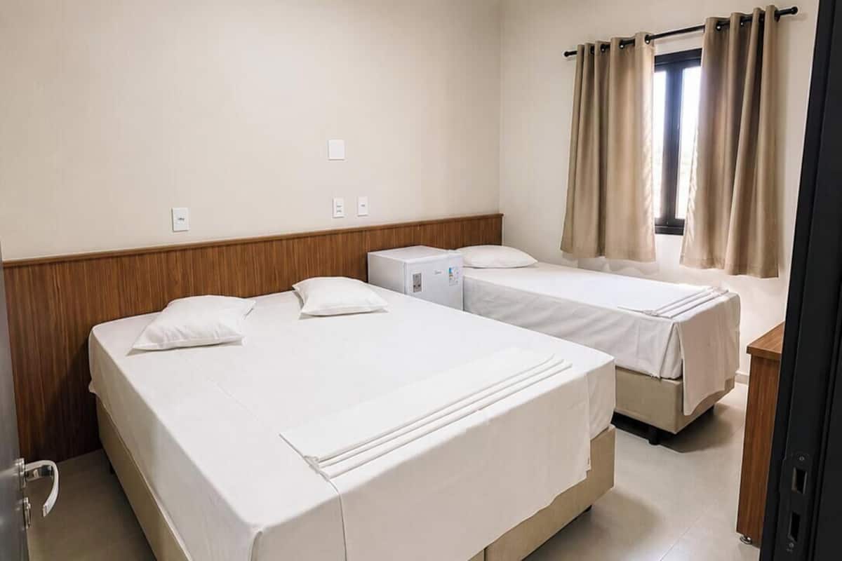 Quarto de hotel com duas camas, cabeceira de madeira, uma escrivaninha pequena e uma janela com cortinas.