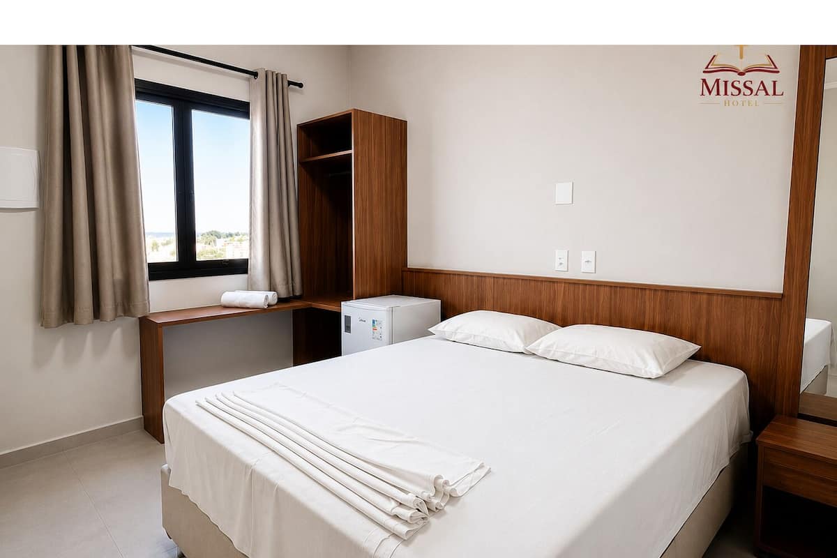 Quarto de hotel com cama, escrivaninha, guarda-roupa e janela com cortinas.