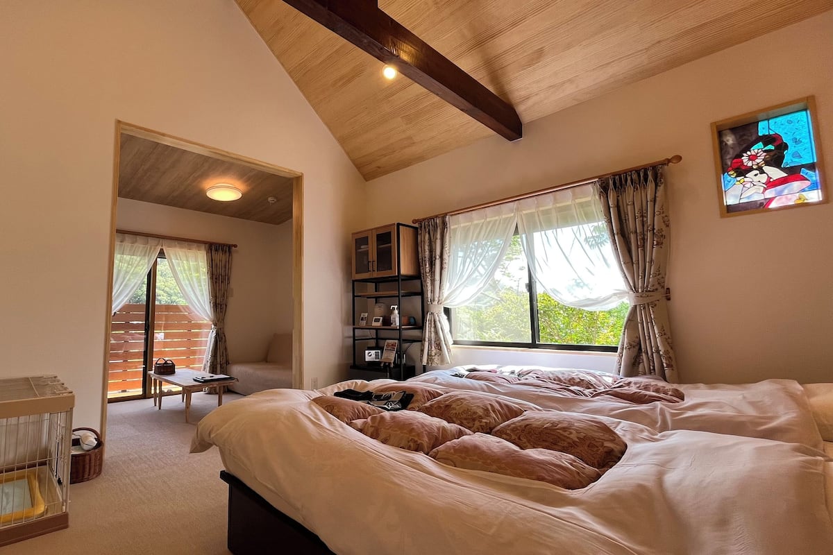 Um quarto com uma cama grande, teto de madeira, uma mesinha e uma janela com cortinas.