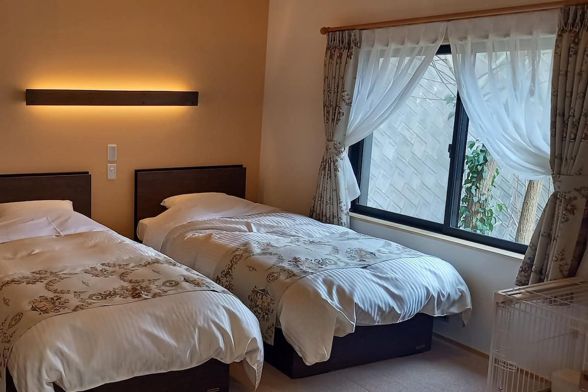 Quarto com duas camas, uma mesinha, uma cadeira, um radiador e janela com cortinas.
