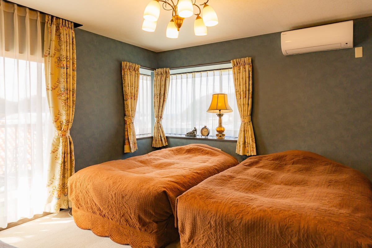 Quarto com duas camas, uma janela com cortinas, uma luminária e um aparelho de ar condicionado.