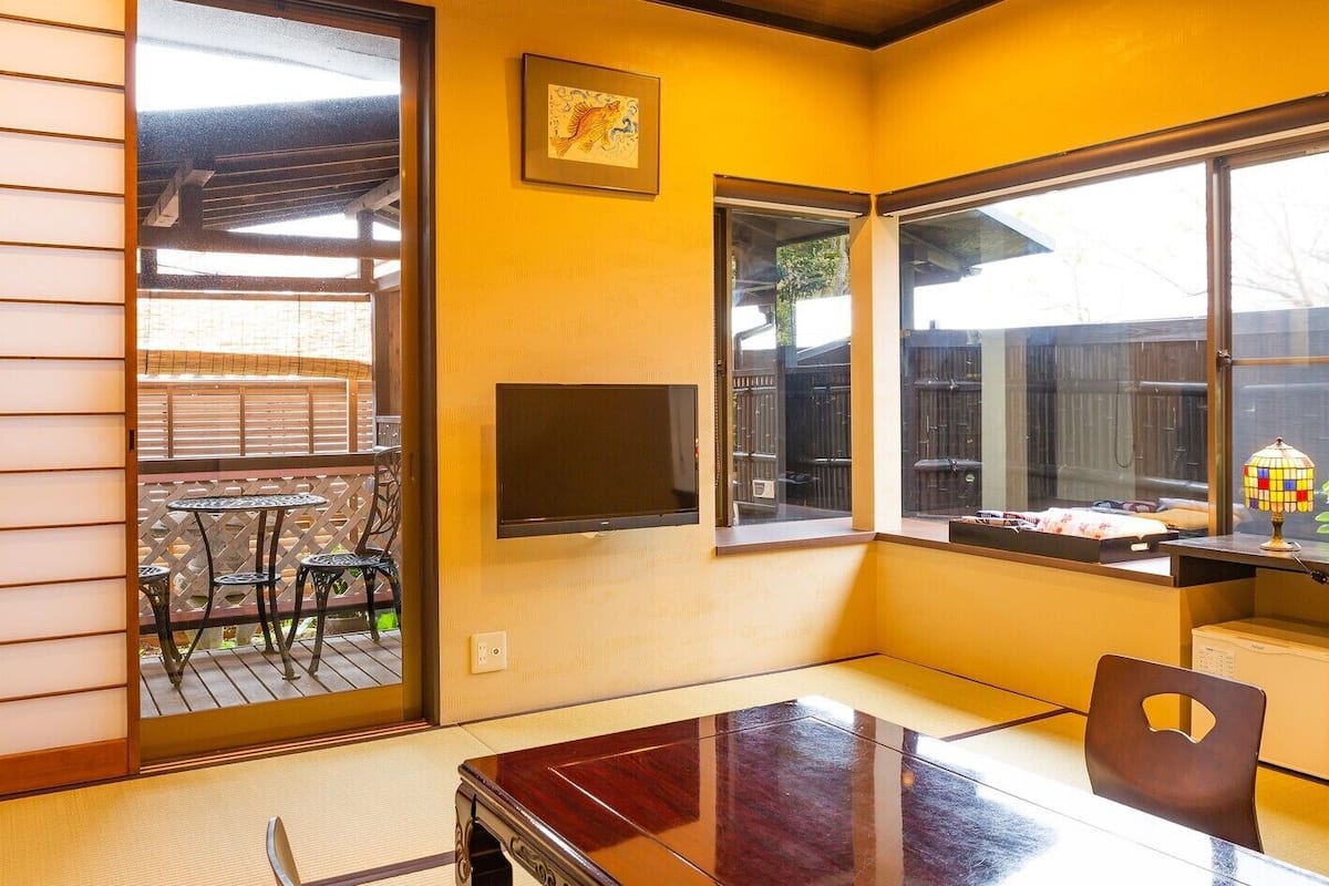 Quarto com piso de tatame, porta deslizante para área externa, televisão fixada na parede e mesa com cadeiras.