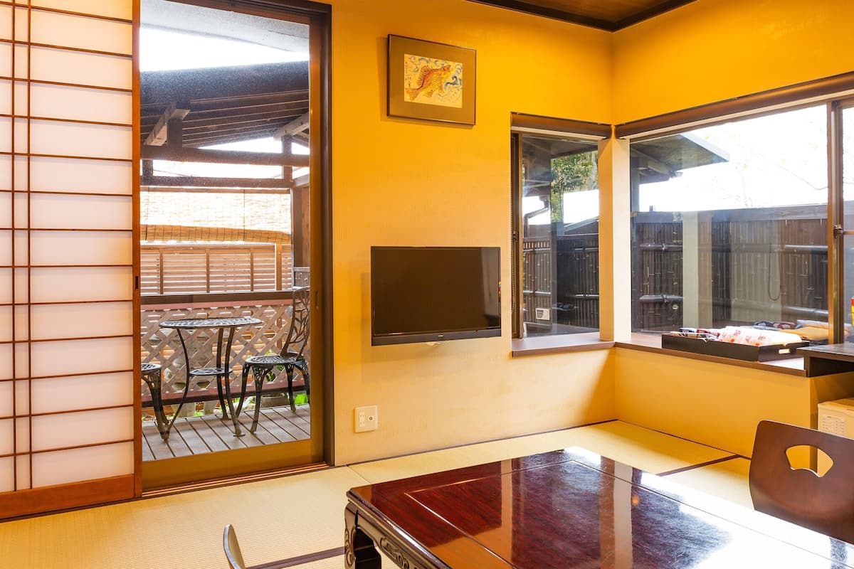 Quarto com piso de esteira de tatame, uma mesa de madeira, televisão fixada na parede e janela com vista para um quintal.