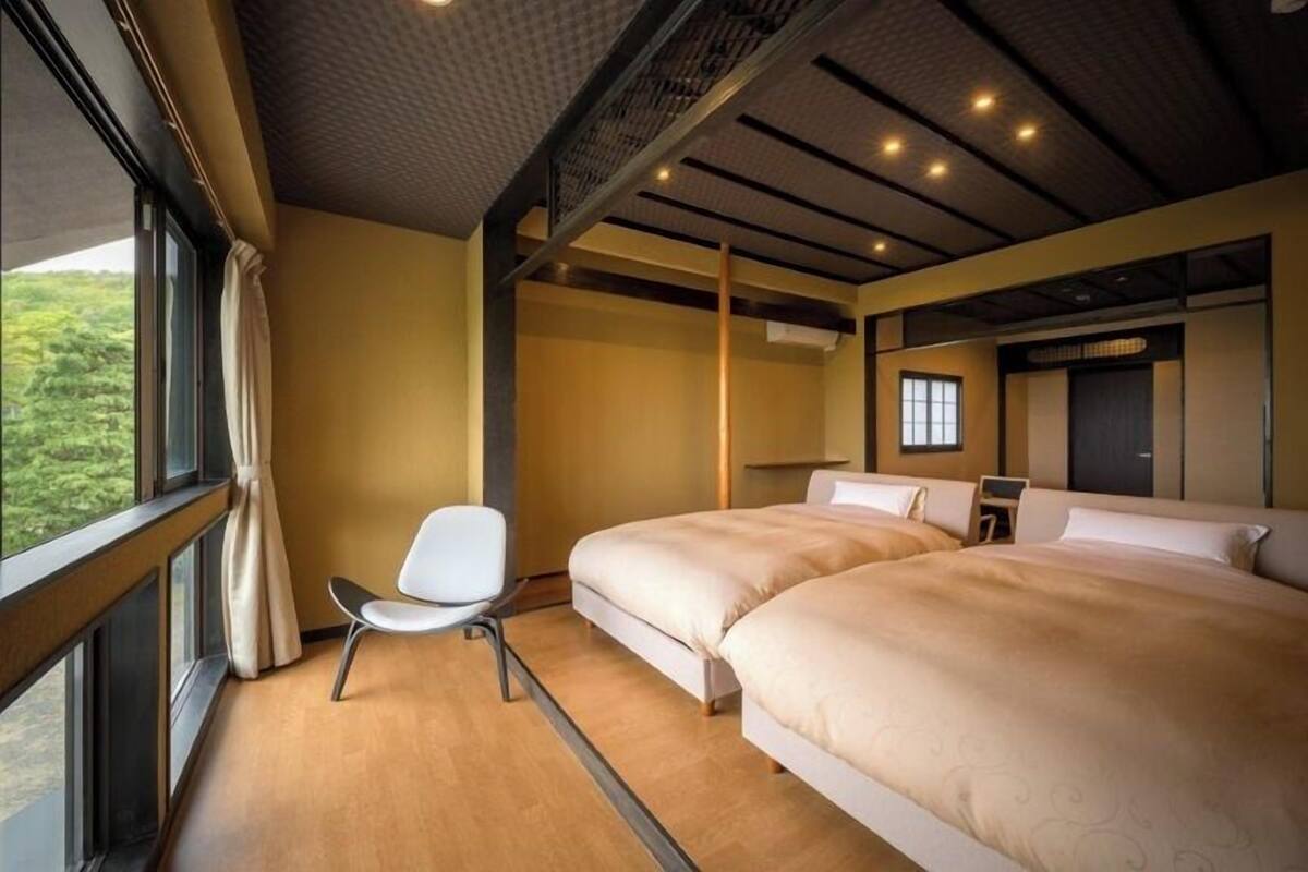 Quarto de hotel com duas camas, uma cadeira, uma janela grande e vista para o verde lá fora.