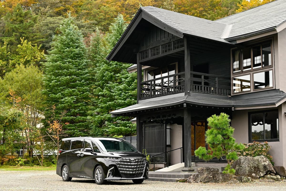 Um edifício de dois andares com varanda, um carro estacionado na frente e um fundo arborizado.