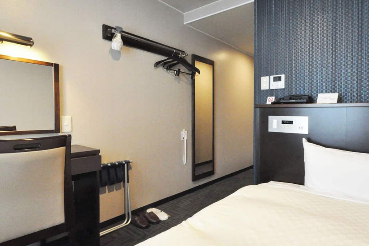 Quarto de hotel com cama, escrivaninha, espelho, cabideiro e telefone.