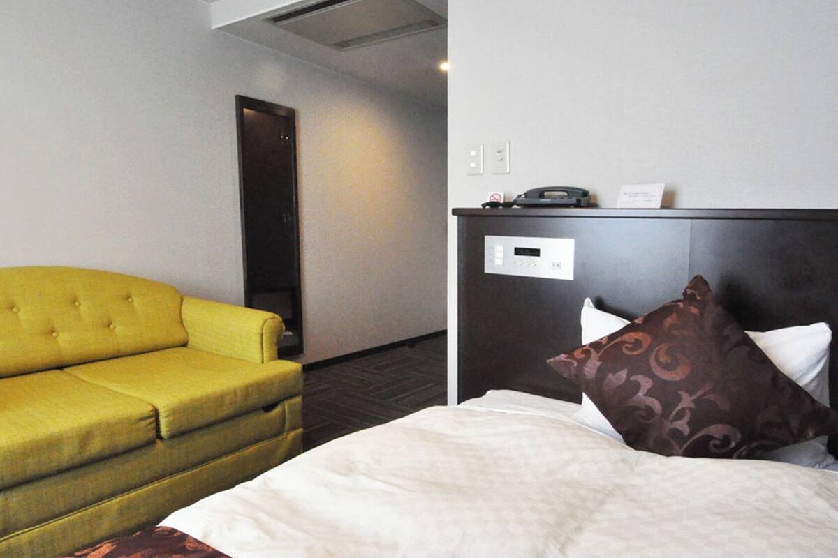 Quarto de hotel com sofá amarelo, cama com edredom branco e almofadas castanhas, cabeceira escura e pequena mesa com telefone e placa.