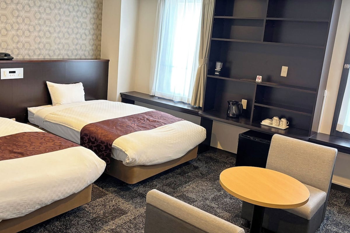 Quarto de hotel com duas camas, uma mesinha, uma cadeira, um minibar e uma janela com cortinas.