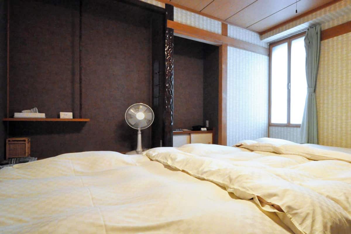 Quarto de hotel com uma cama, um ventilador, uma janela com cortinas e uma estante com objetos.