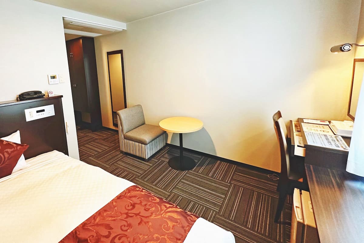 Quarto de hotel com cama, uma cadeira, uma mesinha e uma escrivaninha com telefone.
