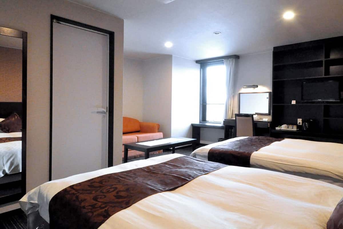 Quarto de hotel com duas camas, uma escrivaninha, uma cadeira, um sofá e uma janela grande.