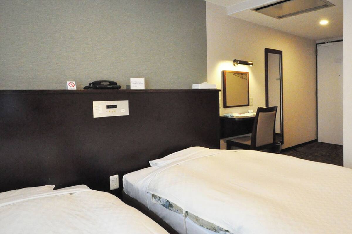 Quarto de hotel com duas camas, uma escrivaninha, uma cadeira, um espelho e uma porta.