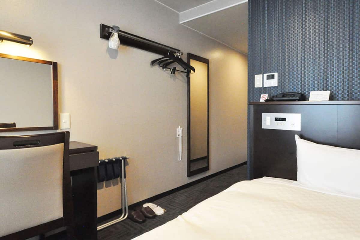 Quarto de hotel com cama, escrivaninha, espelho, cabideiro, telefone e painel de controle fixado na parede.