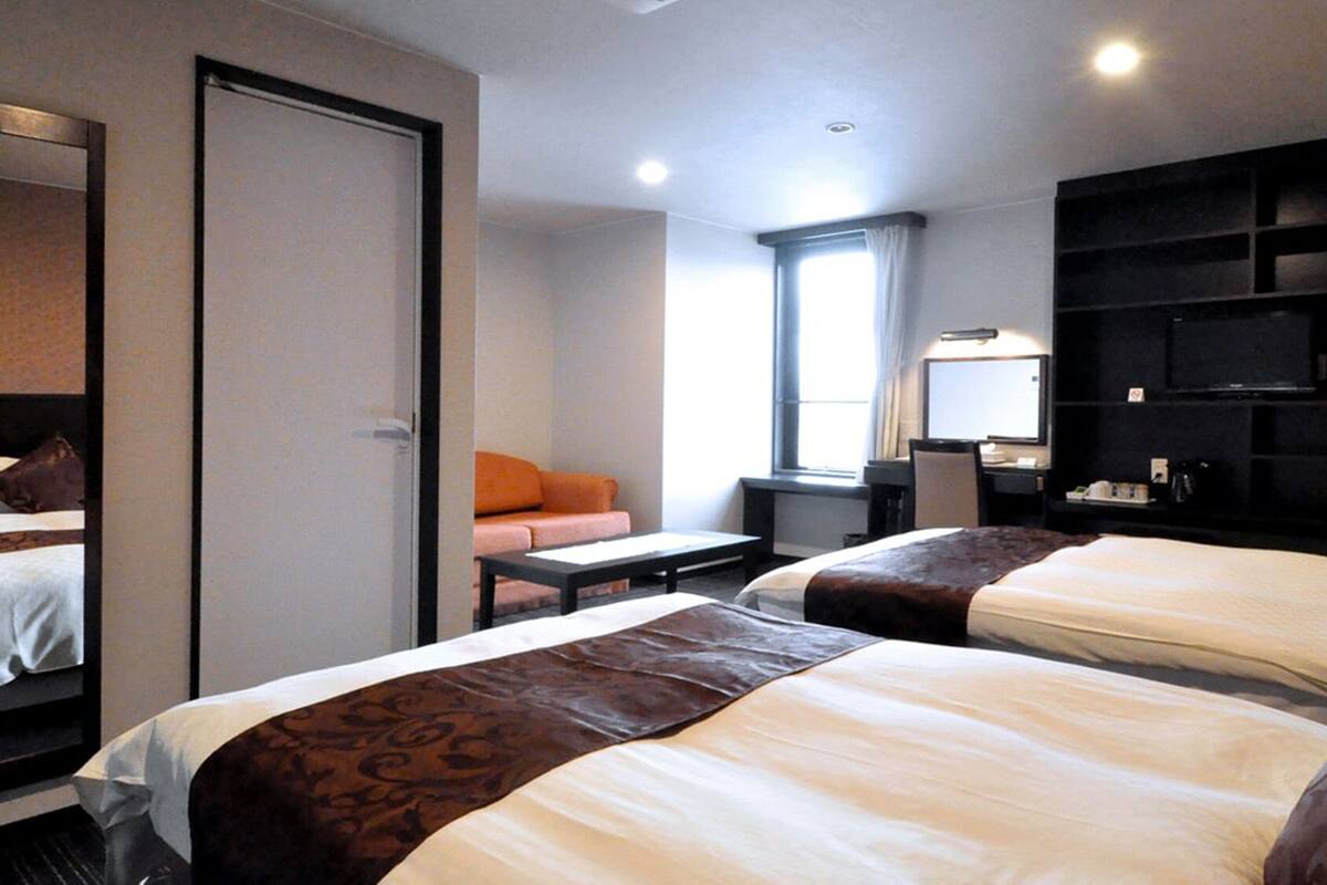 Quarto de hotel com duas camas, um sofá, uma escrivaninha e uma janela ampla.