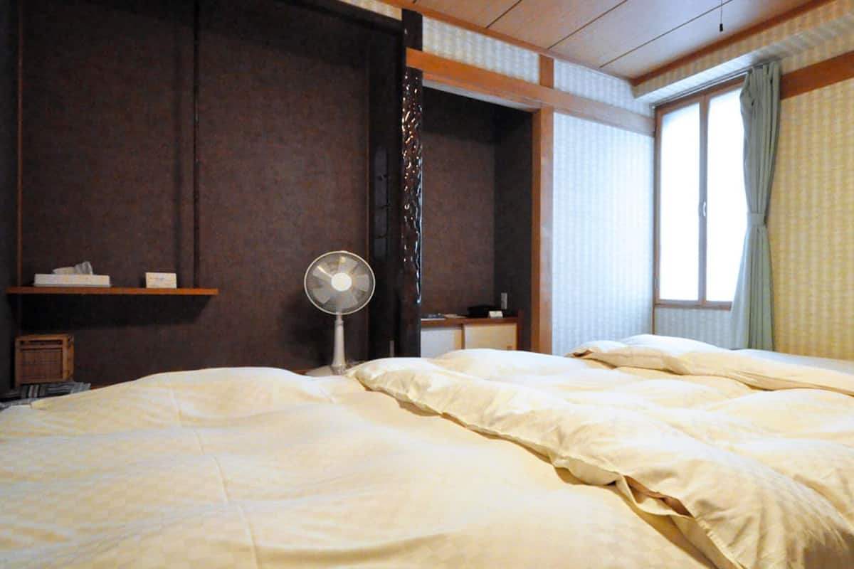 Quarto de hotel com cama, ventilador, janela com cortinas e prateleira com objetos.