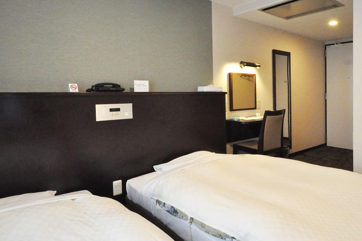 Quarto de hotel com duas camas, uma escrivaninha, uma cadeira, uma luminária de mesa, um espelho e uma porta.