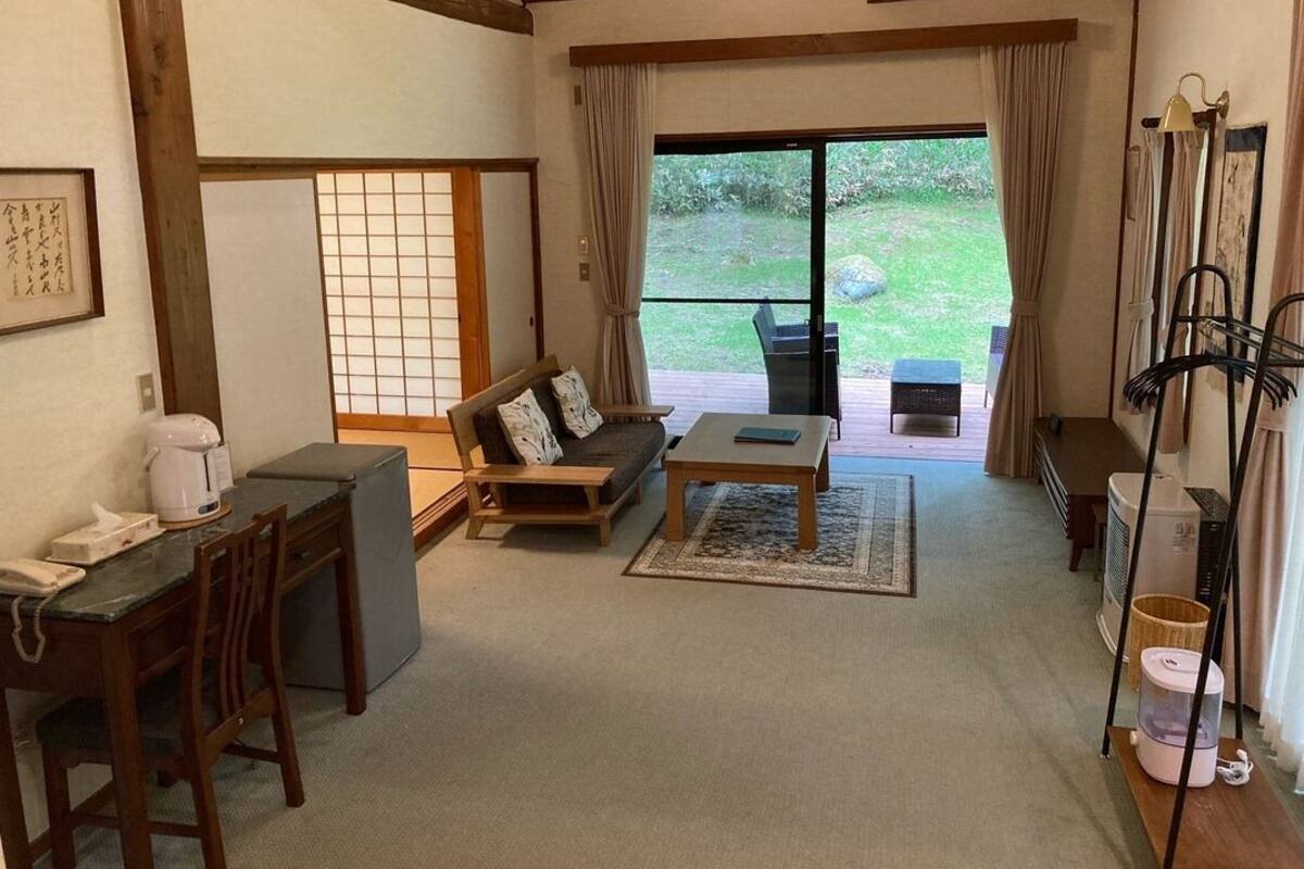 Quarto em estilo japonês tradicional com tatames, uma mesa baixa, um sofá, uma porta de vidro deslizante para um jardim e uma pequena mesa com um chuveiro elétrico.