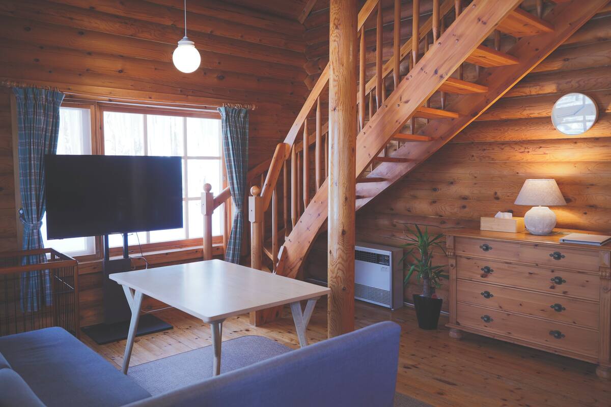 Interior acolhedor de uma cabana, com escada de madeira, TV de tela plana, mesa de jantar, sofá e cómoda.