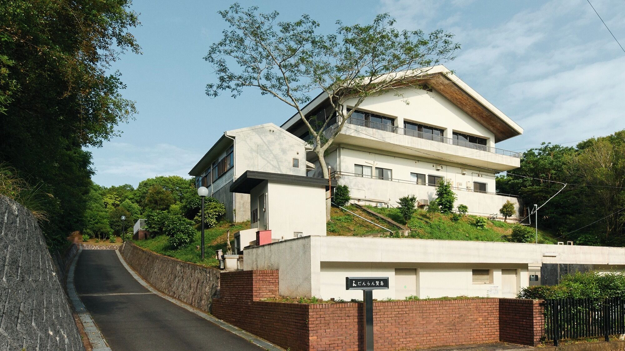 Um edifício moderno de dois andares com telhado inclinado, uma escadaria visível que leva ao andar superior e uma placa de rua indicando o nome do local.