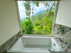 Casa de banho com banheira, chuveiro e janela com vista para uma floresta e montanhas.