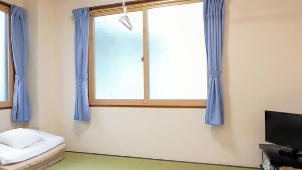 Quarto com piso de tatame, cama com roupa de cama branca, televisão em um suporte e cortinas azuis nas janelas.