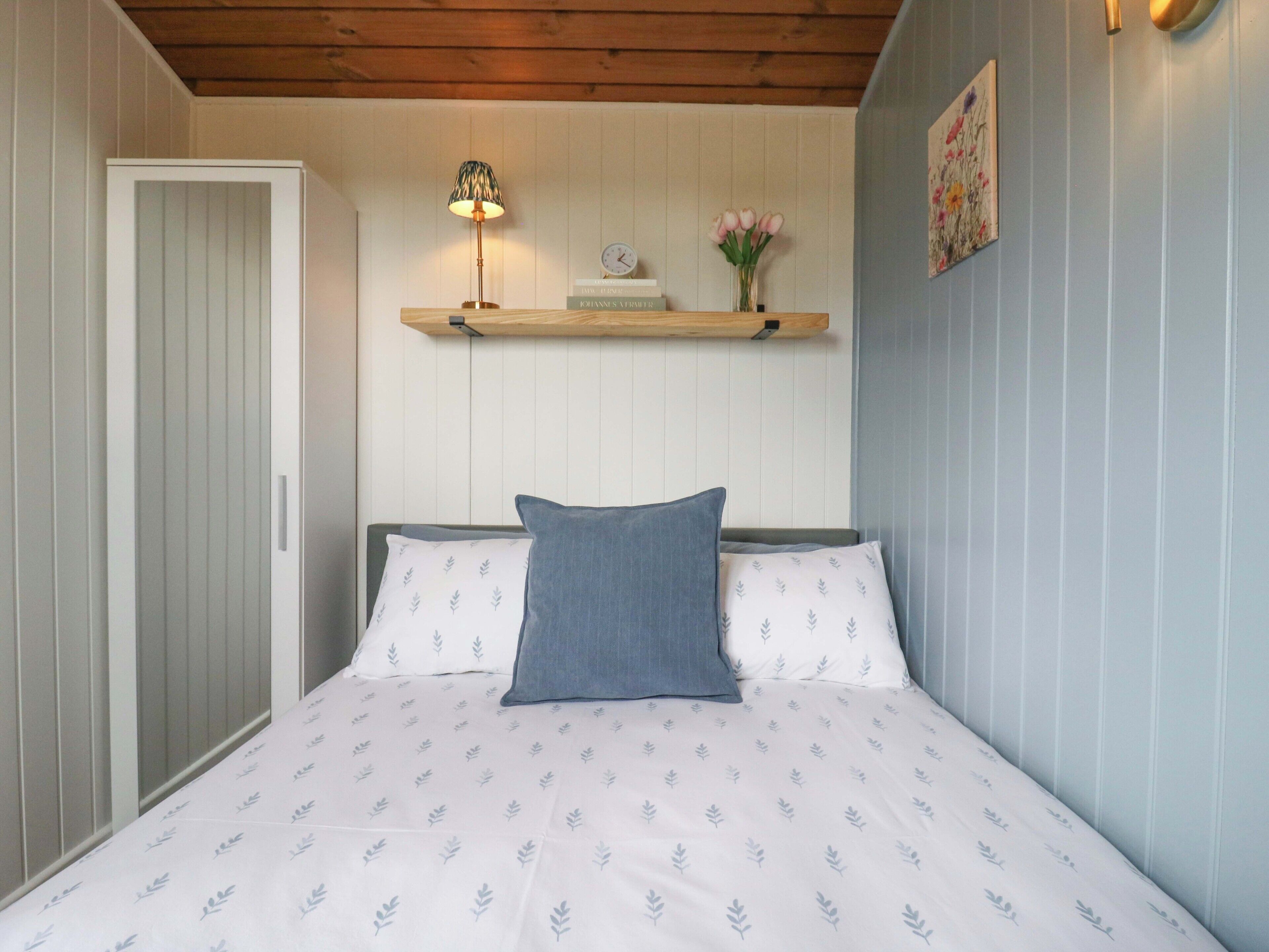 Quarto com cama, mesa de cabeceira, prateleira na parede com luminária, relógio e vaso com flores.