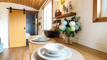 Interior de madeira com área de refeições, mesa posta com pratos e tigelas brancos, um vaso com flores e uma prateleira fixada na parede com objetos decorativos.