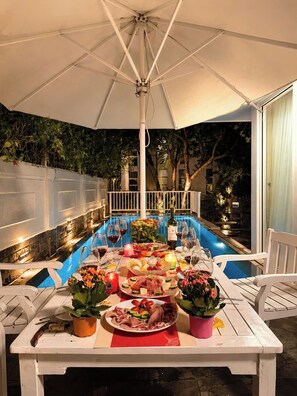 Uma área de refeições ao ar livre com piscina, mobiliário branco e uma mesa posta para refeição.