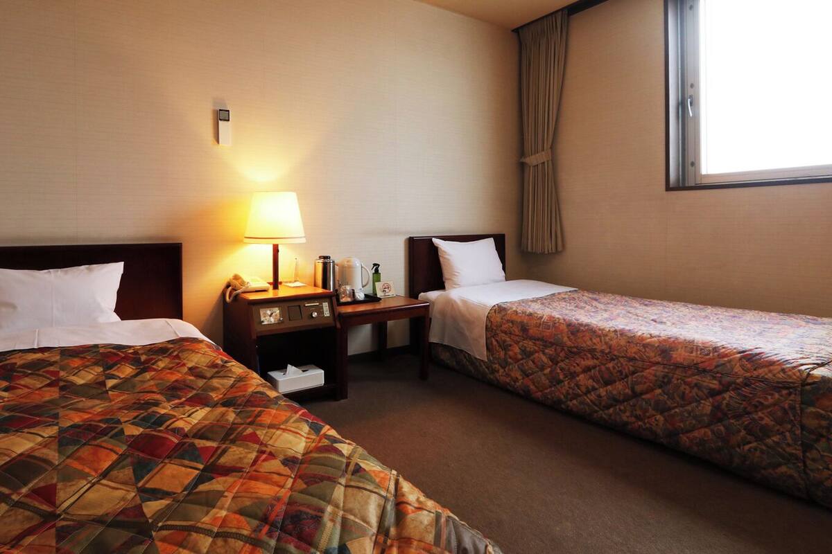 Quarto de hotel com duas camas, uma mesinha de cabeceira com abajur, telefone e uma pequena mesa com garrafa e caixa de lenços.