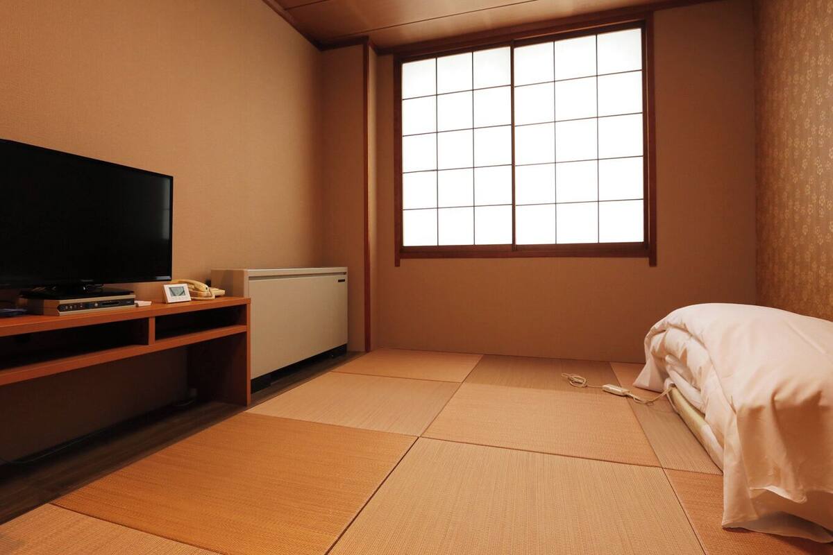 Quarto com piso de tatame, televisão em suporte de madeira, cama com roupa de cama branca e janela com padrão de grade.