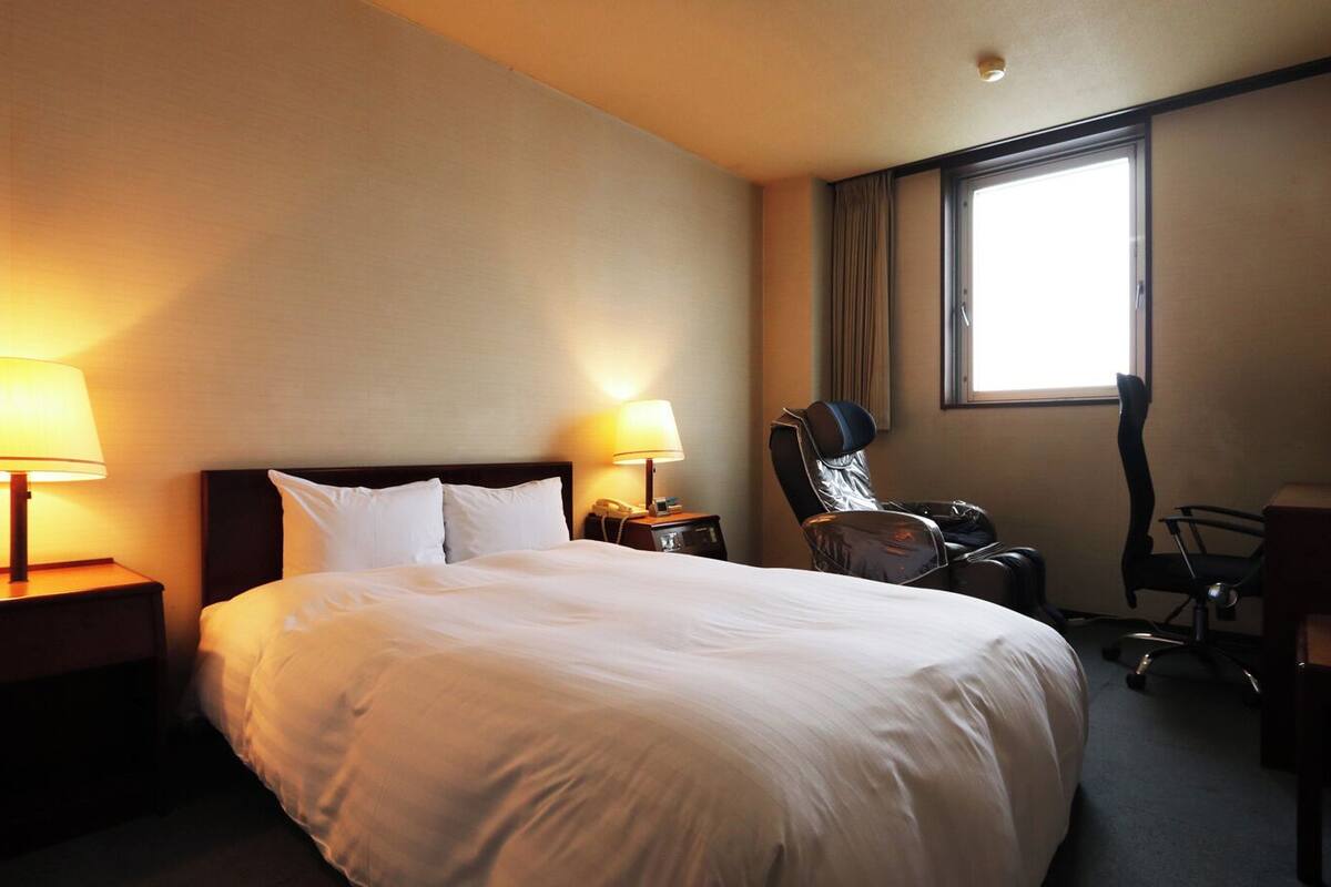 Quarto de hotel com uma cama grande, duas luminárias de cabeceira, uma cadeira de massagem, uma escrivaninha com cadeira e uma janela.