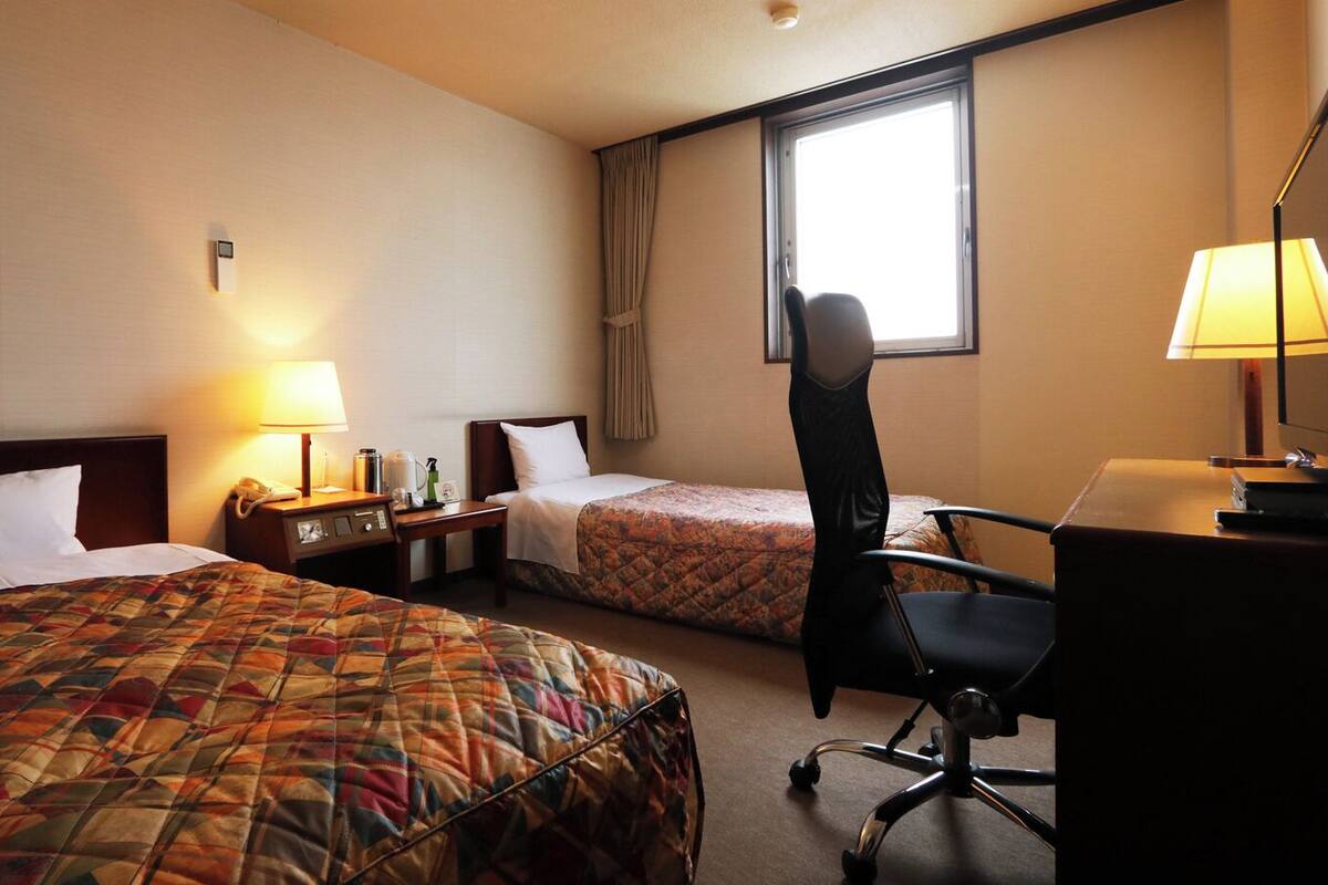 Quarto de hotel com duas camas, uma escrivaninha, uma cadeira, uma janela e um abajur.