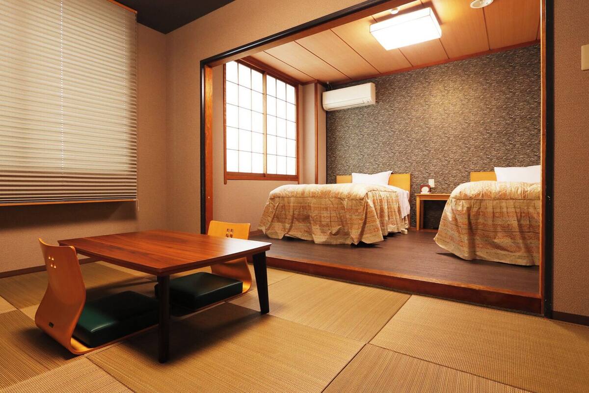 Quarto com piso de tatame, uma mesa de madeira, duas camas com roupa de cama estampada e uma porta deslizante para outro quarto.