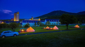 Um hotel com várias tendas montadas num campo gramado, um edifício grande com uma torre peculiar e uma montanha ao fundo.