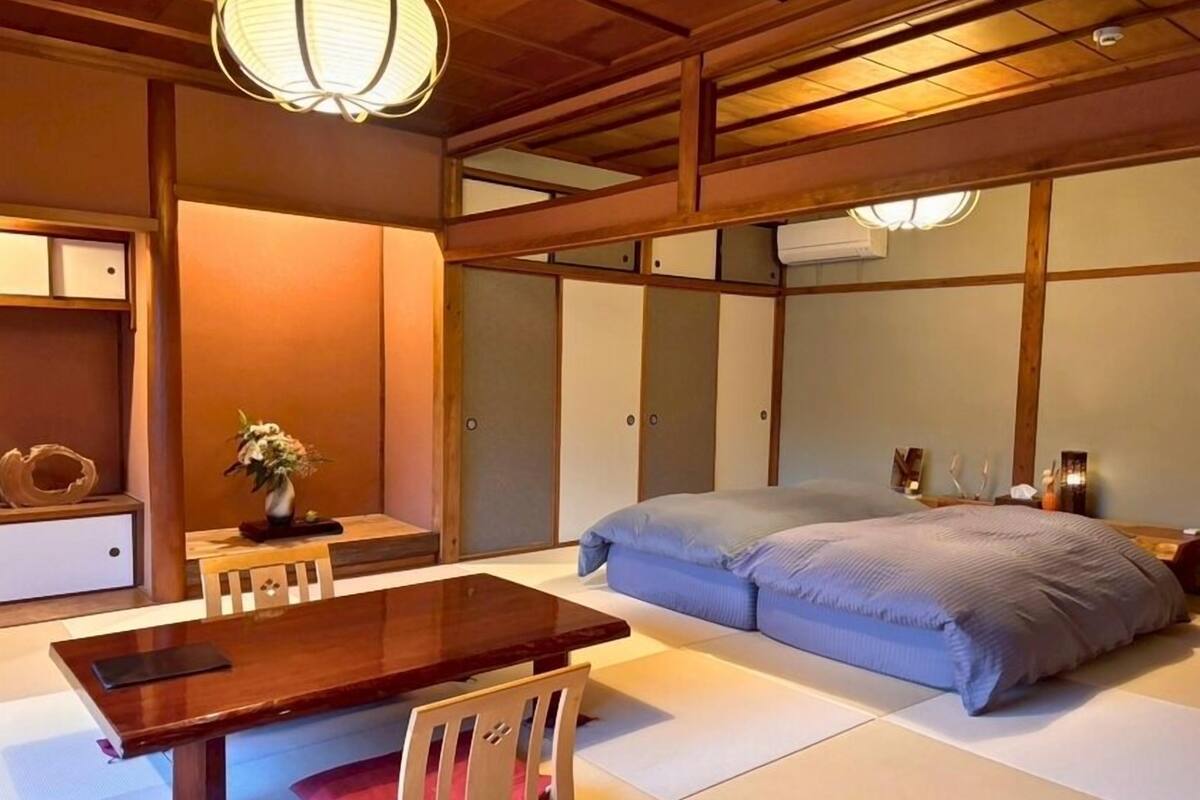Um quarto tradicional em estilo japonês, com piso de tatame, uma mesa de madeira baixa, uma cama com roupa de cama azul e um lanterna de papel pendurada.