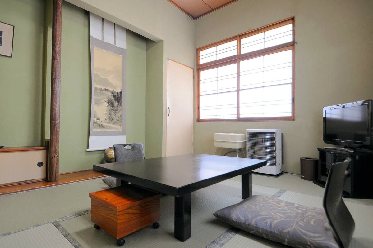 Quarto com piso de esteira de tatame, mesa baixa com almofada, televisão e uma janela grande com persianas.