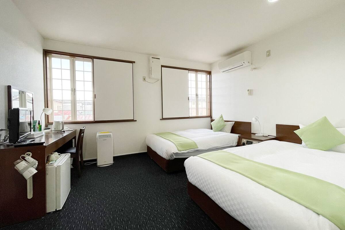 Quarto de hotel com duas camas, uma escrivaninha com computador, uma cadeira, televisão e uma janela.
