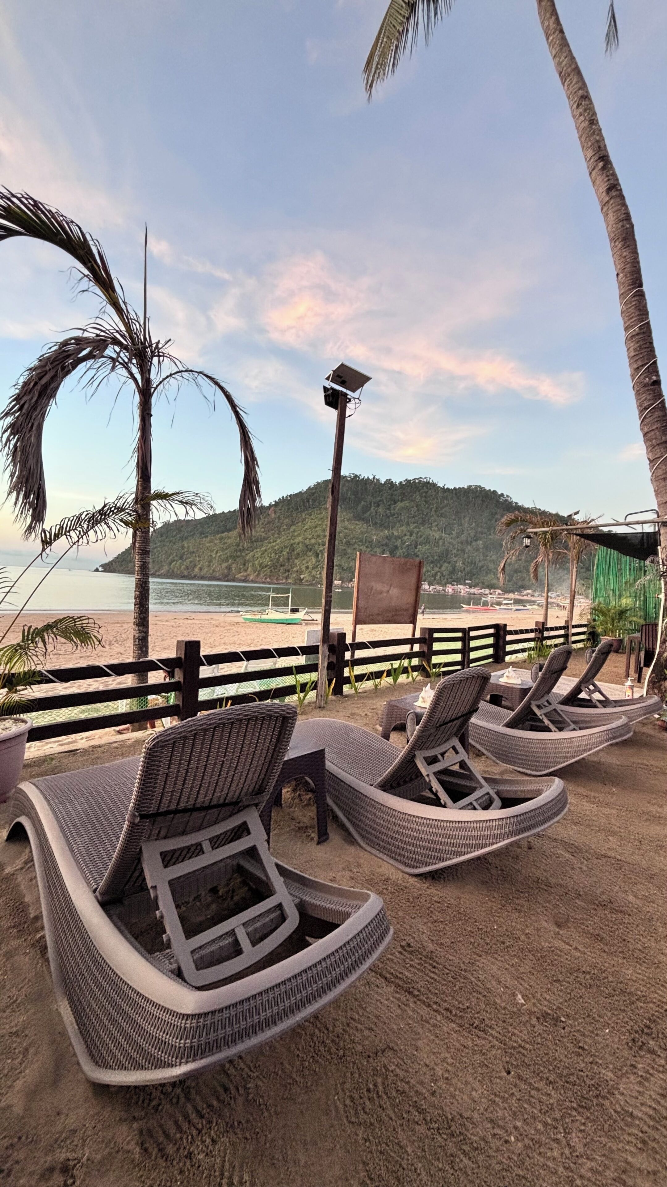 Cadeiras de espreguiçadeira de vime em uma praia arenosa, com vista para o mar e uma montanha ao fundo.